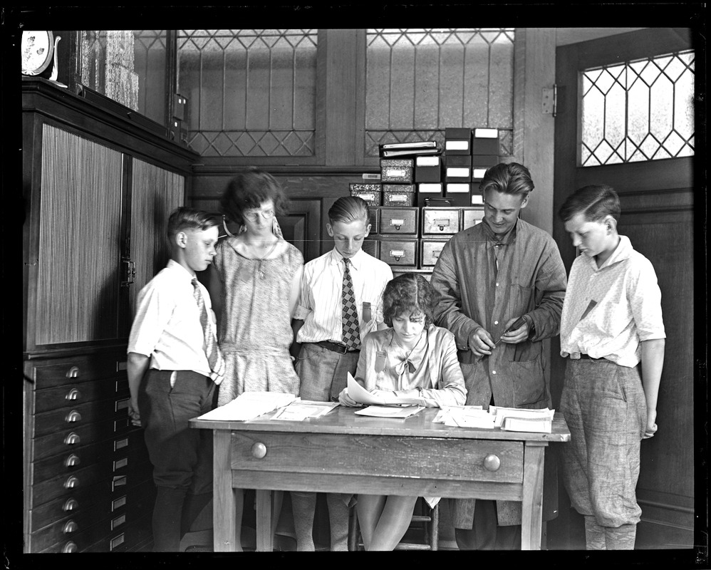 Child sits at desk reading papers while children stand behind her and ...