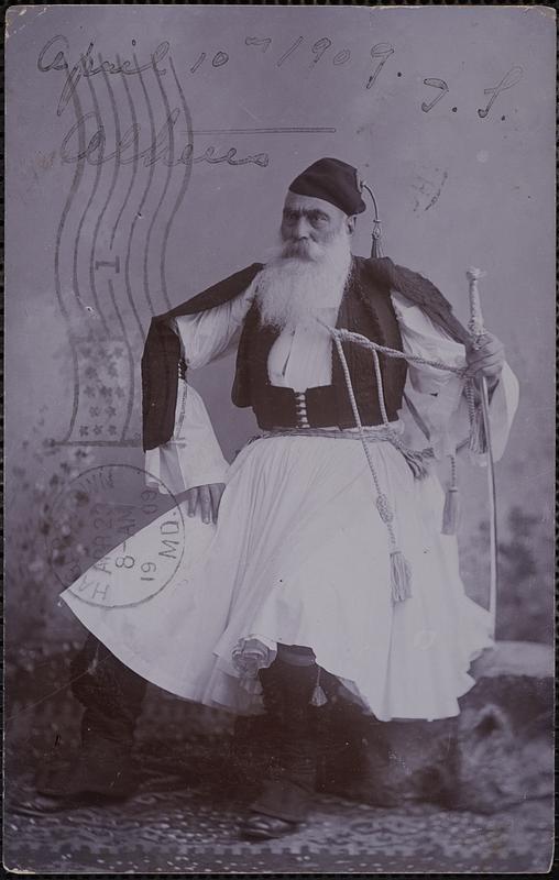 A bearded man sits wearing traditional Greek dress including a ...