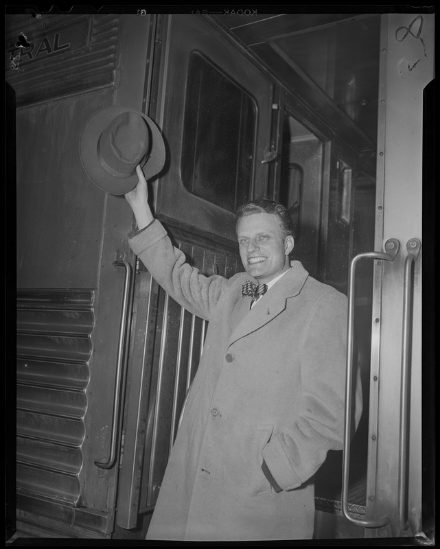 Billy Graham standing near a train car with hat raised in hand ...