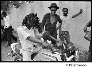 Dillinger (reggae musician), seated on a motorcycle, tilting his head ...
